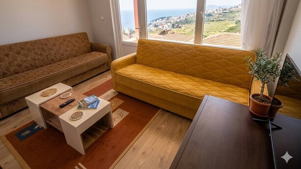 Uma sala de estar com um sofá marrom, um sofá amarelo, uma mesa de centro e vista para uma paisagem costeira através de grandes janelas.
