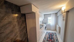 Um corredor estreito com piso de azulejos, uma área de cozinha com armários brancos e um fogão, e uma janela com cortinas.