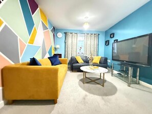 Uma sala de estar com um sofá amarelo, um sofá preto, uma mesa de centro branca e uma televisão sobre um aparador de vidro.