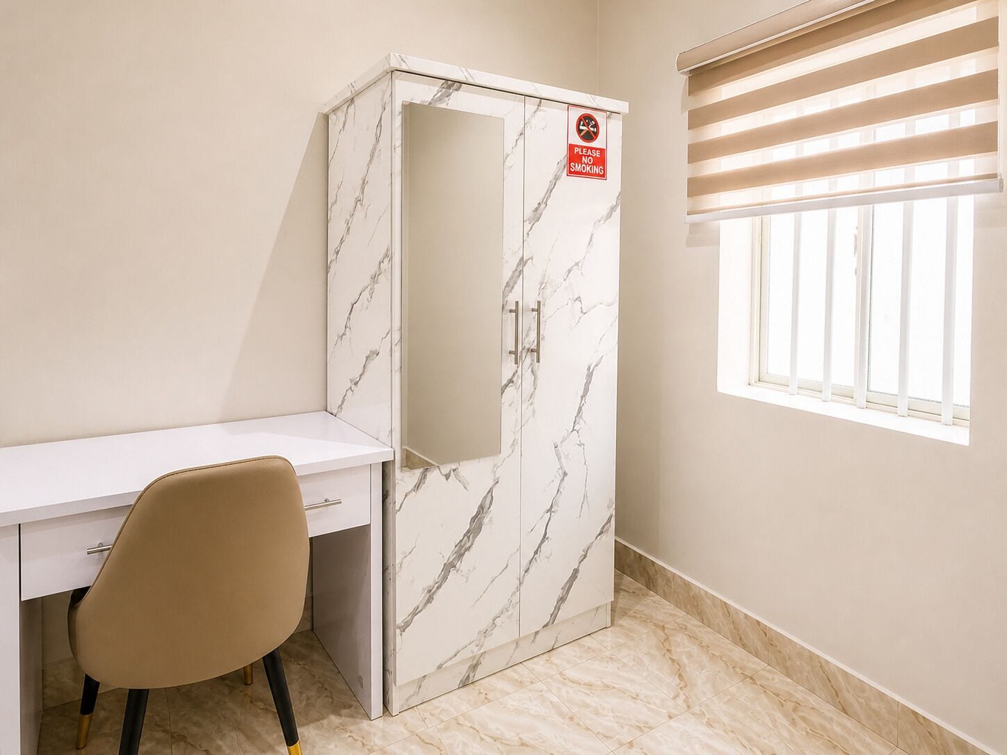 Um quarto com uma secretária branca, uma cadeira bege, um guarda-roupa com padrão marmorizado e uma janela com persianas.