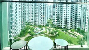 Uma varanda com quatro mesas redondas e guarda-corpo de vidro, com vista para uma área residencial com edifícios altos e um parque.