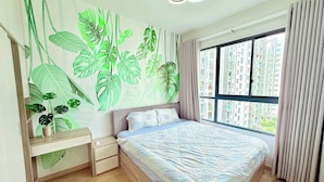 Um quarto com uma cama grande, uma mesa de cabeceira, um mural de folhas verdes na parede, uma janela com cortinas e vista para edifícios lá fora.