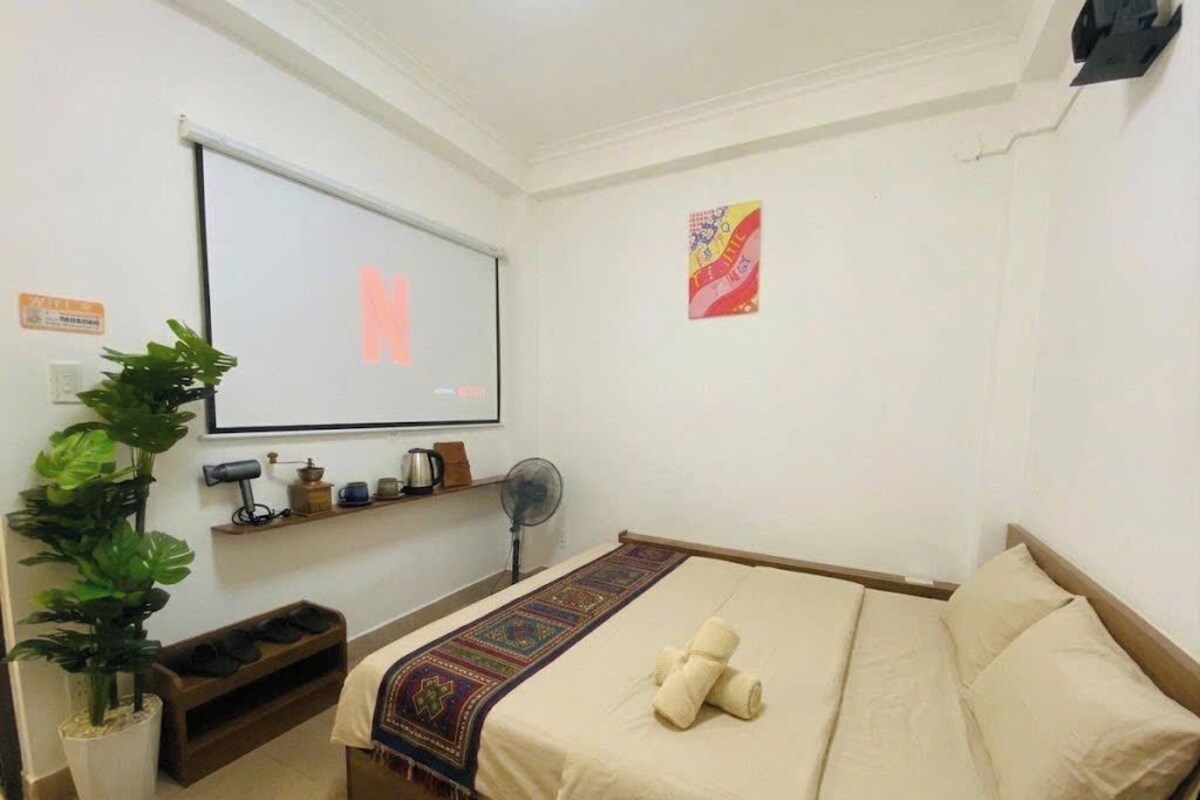 Quarto com cama, tela de projetor, ventilador, prateleira com objetos e uma planta.