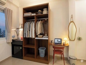 Quarto compacto com guarda-roupa de madeira, escrivaninha com laptop, cadeira, lixeira pequena e espelho na parede.