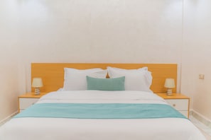 Cama bem arrumada com cabeceira de madeira, duas mesas de cabeceira com luminárias e um manta azul-esverdeada.