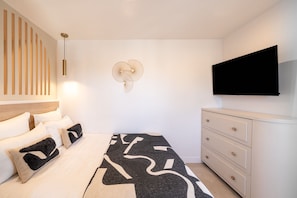 Quarto com cama, almofadas, mesa de cabeceira, cômoda, televisão e luminária fixada na parede.