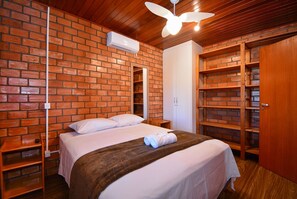 Um quarto com parede de tijolos, cama com roupa de cama branca, ventilador de teto e prateleiras de madeira embutidas.