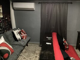 Quarto com sofá cinza, um sofá preto e vermelho, uma mesinha com controle remoto e uma parede cinza com janela.
