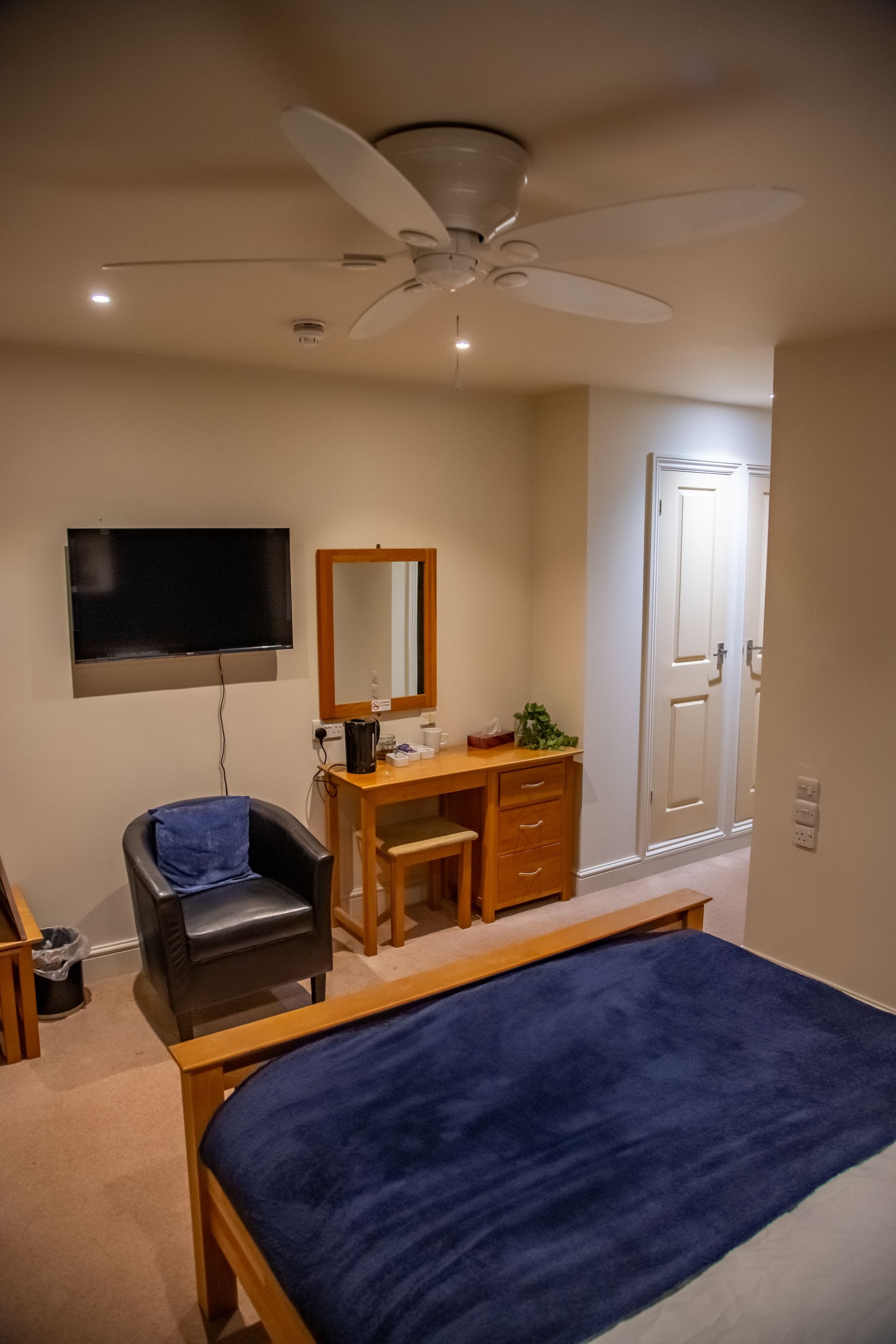 Quarto com cama, escrivaninha, cadeira, televisão, ventoinha no teto e porta.