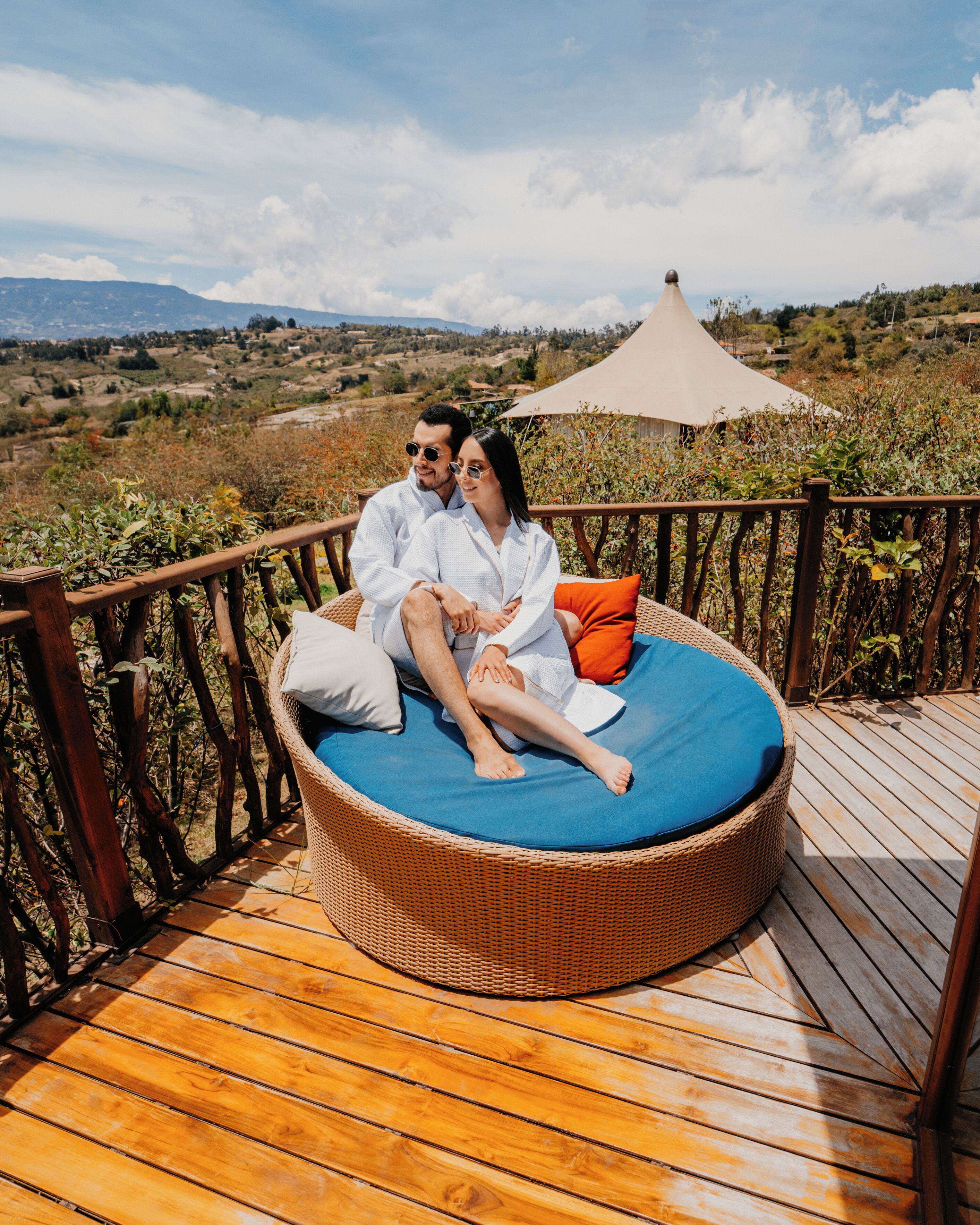 Um casal sentado em uma cadeira de vime com almofada azul em um deck de madeira, com vista para uma paisagem montanhosa.