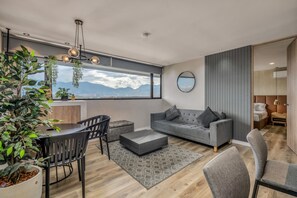 Uma sala de estar moderna com um sofá cinzento, uma mesa de jantar com cadeiras, uma janela ampla com vista para montanhas e um lustre.