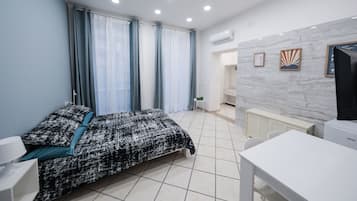 Quarto com cama, escrivaninha e cadeira. Há uma janela com cortinas, um radiador e ar condicionado fixado na parede.
