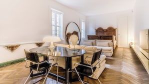 Uma área de refeições com uma mesa redonda, quatro cadeiras e uma luminária. Há um sofá e um armário de madeira ao fundo.