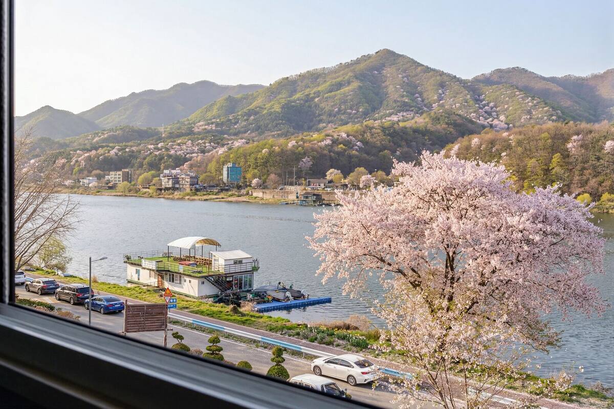Vista de um lago com montanhas ao fundo, uma cerejeira em primeiro plano e um edifício com telhado verde do outro lado da água.