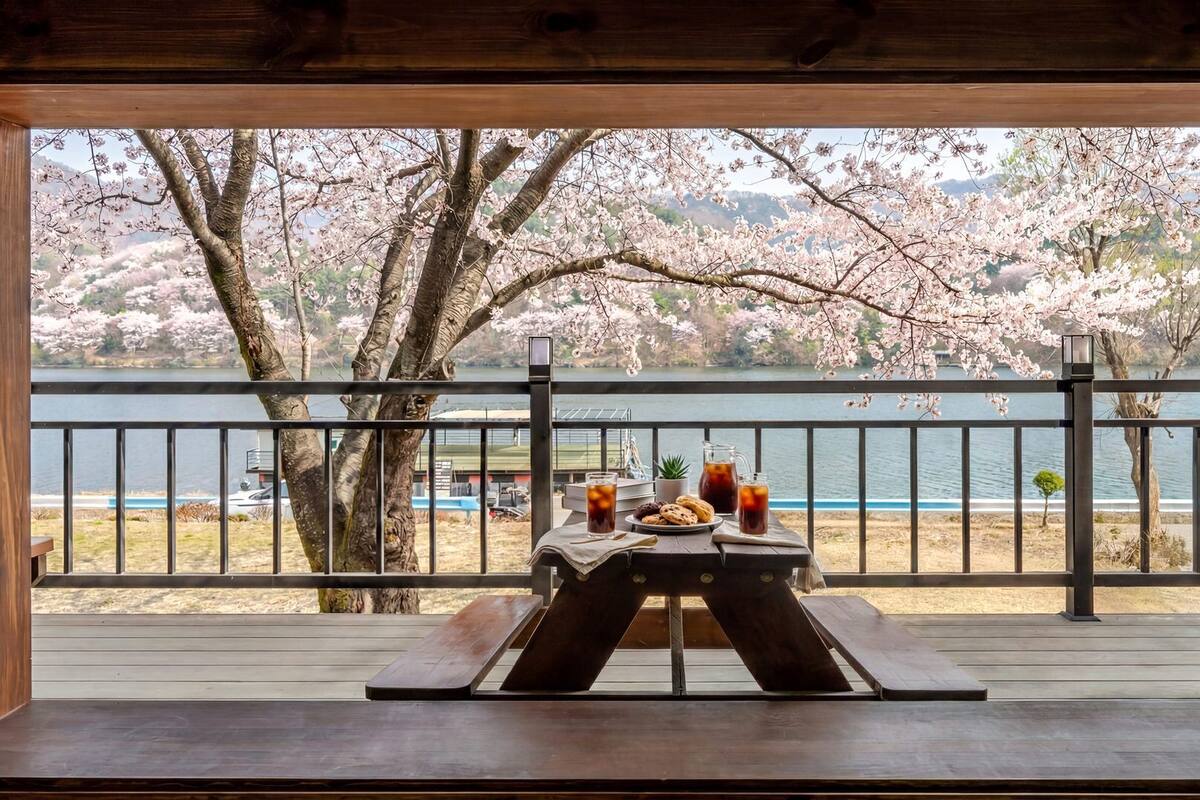 Um deck de madeira com uma mesa de piquenique, vista para um lago, cerejeiras em flor e uma linha de costa distante.