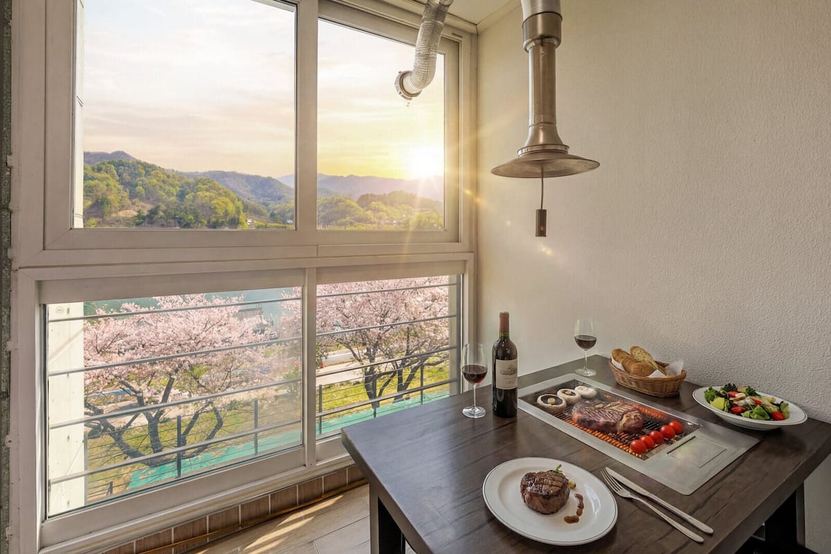 Uma área de refeições com uma mesa posta para uma refeição, uma garrafa de vinho e vista para flores de cerejeira pela janela.