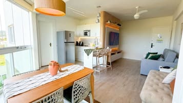 Uma cozinha moderna com armários brancos, micro-ondas e máquina de café. Há uma área de jantar com mesa e cadeiras de madeira. Um sofá cinza está na sala de estar.