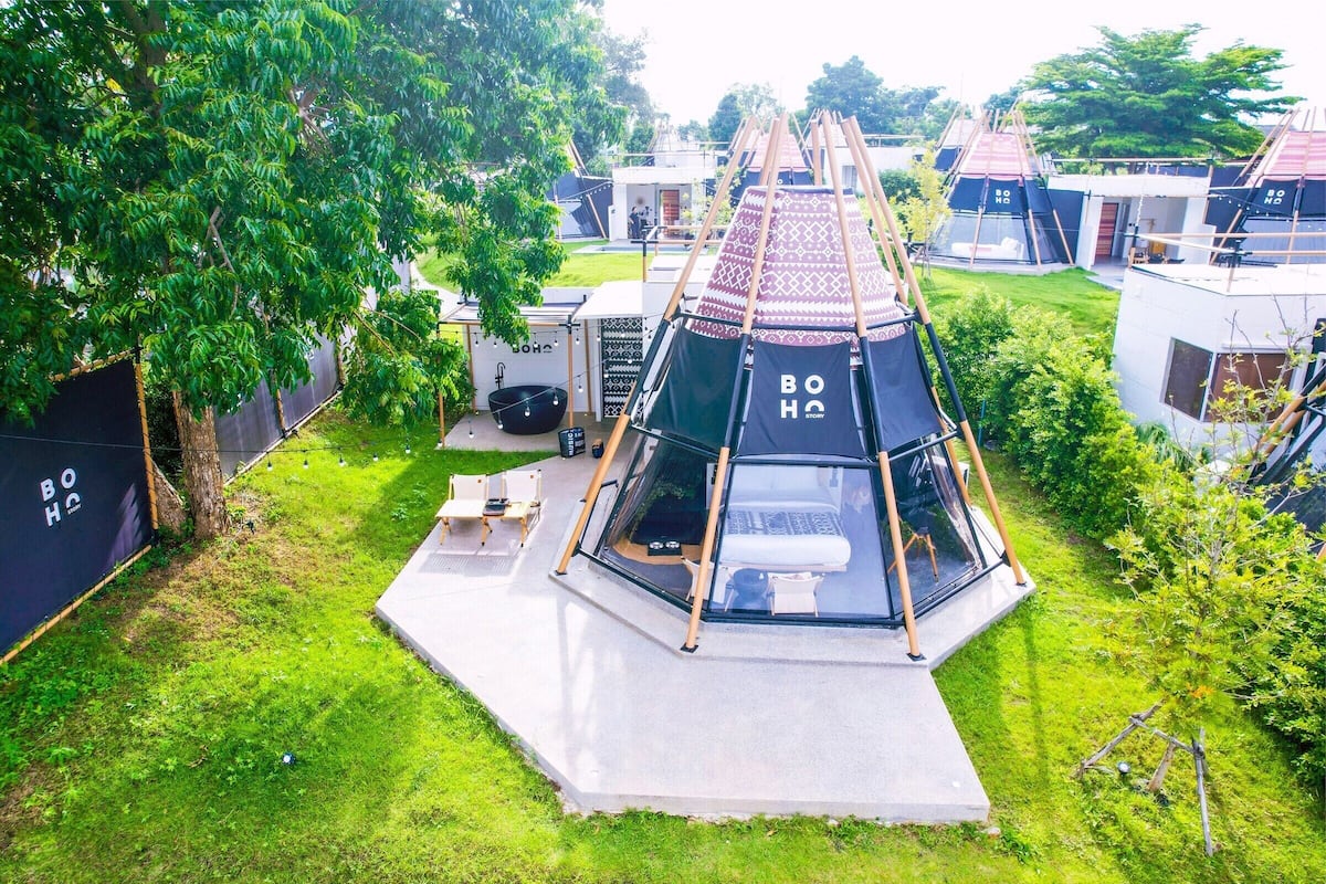 Uma tenda moderna de estilo geométrico com estrutura de vidro, rodeada por uma área gramada e outras tendas semelhantes.