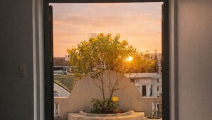 Corredor interno que dá para área externa, com vista para um pôr do sol, uma árvore e um edifício ao fundo.