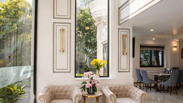 Um quarto amplo com uma grande janela em arco, duas poltronas, uma pequena mesa redonda com flores e uma área de jantar ao fundo.