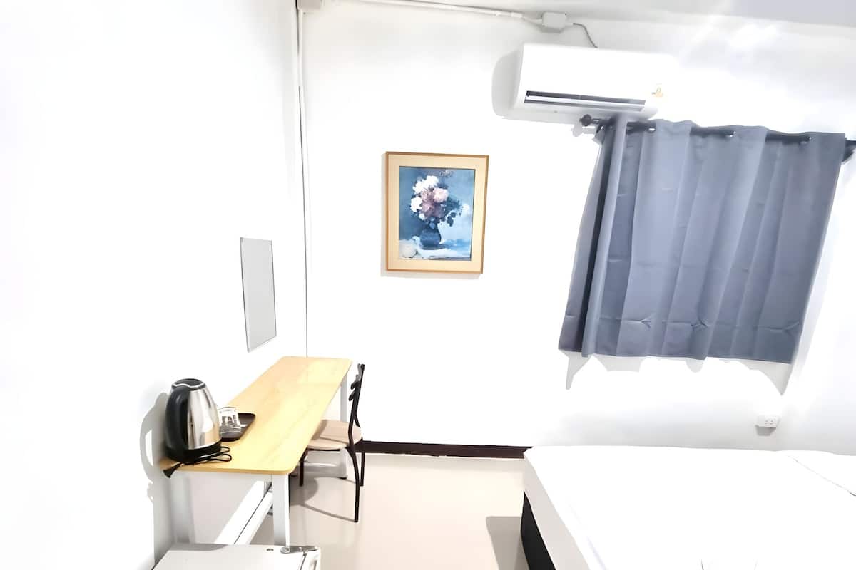 Quarto de hotel compacto com cama, secretária com chaleira, cadeira, mesa pequena, quadro pendurado na parede e janela com cortinas.
