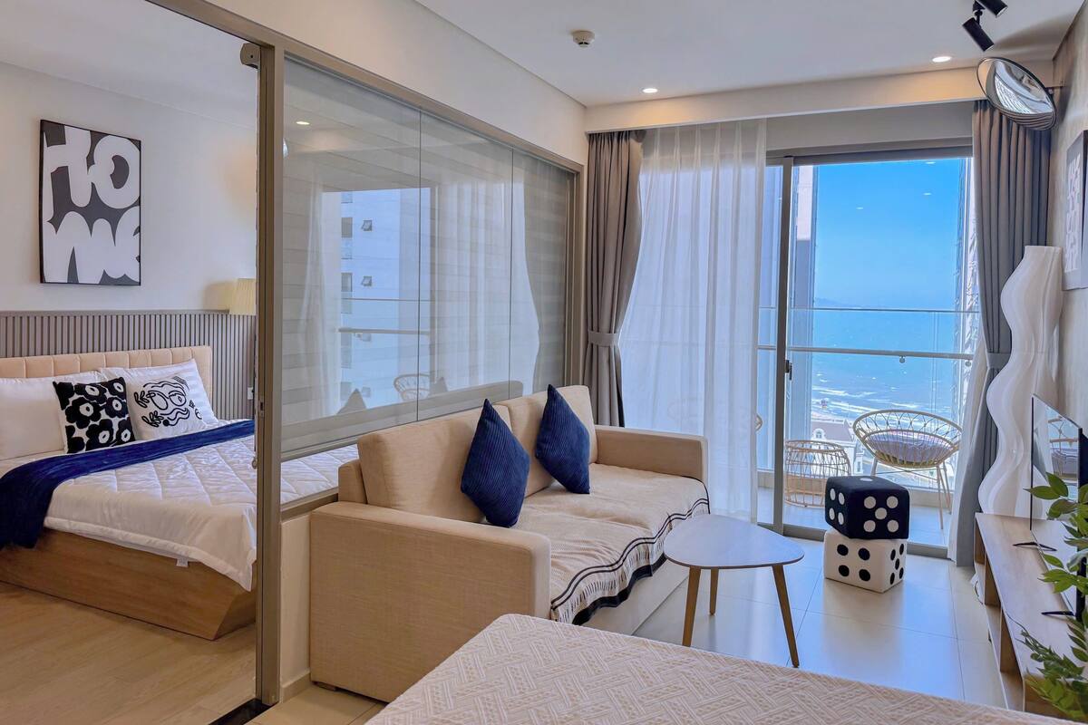 Quarto de hotel moderno com cama, sofá e uma pequena mesa. Há uma grande janela com vista para a cidade.