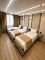 Quarto de hotel com três camas, piso de madeira e uma janela grande com cortinas.