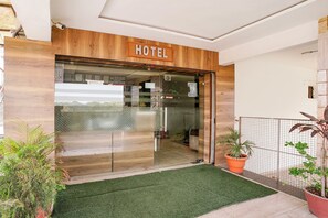 Entrada de hotel com fachada de madeira, porta de vidro e tapete de grama sintética.