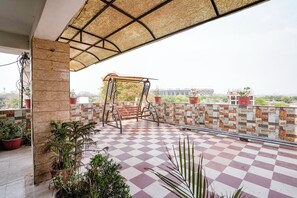Um terraço no topo do prédio com piso xadrez, balanço, plantas em vasos e vista para a paisagem urbana.