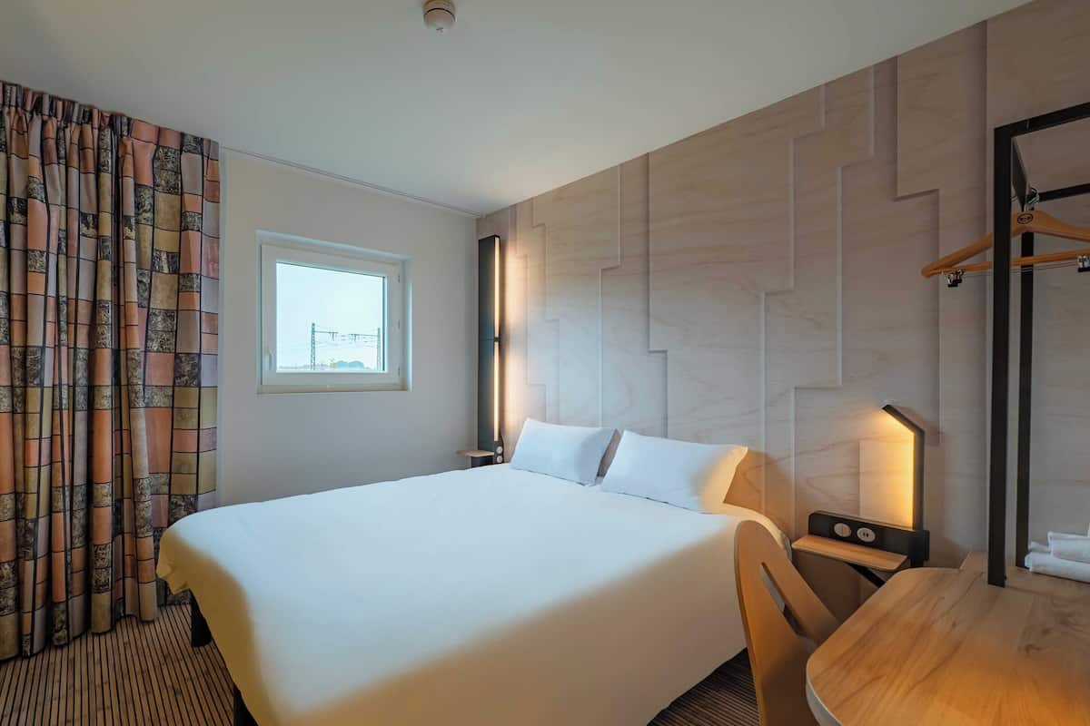 Quarto de hotel com uma cama grande, uma mesa pequena, uma cadeira, uma janela e um candeeiro fixado na parede.