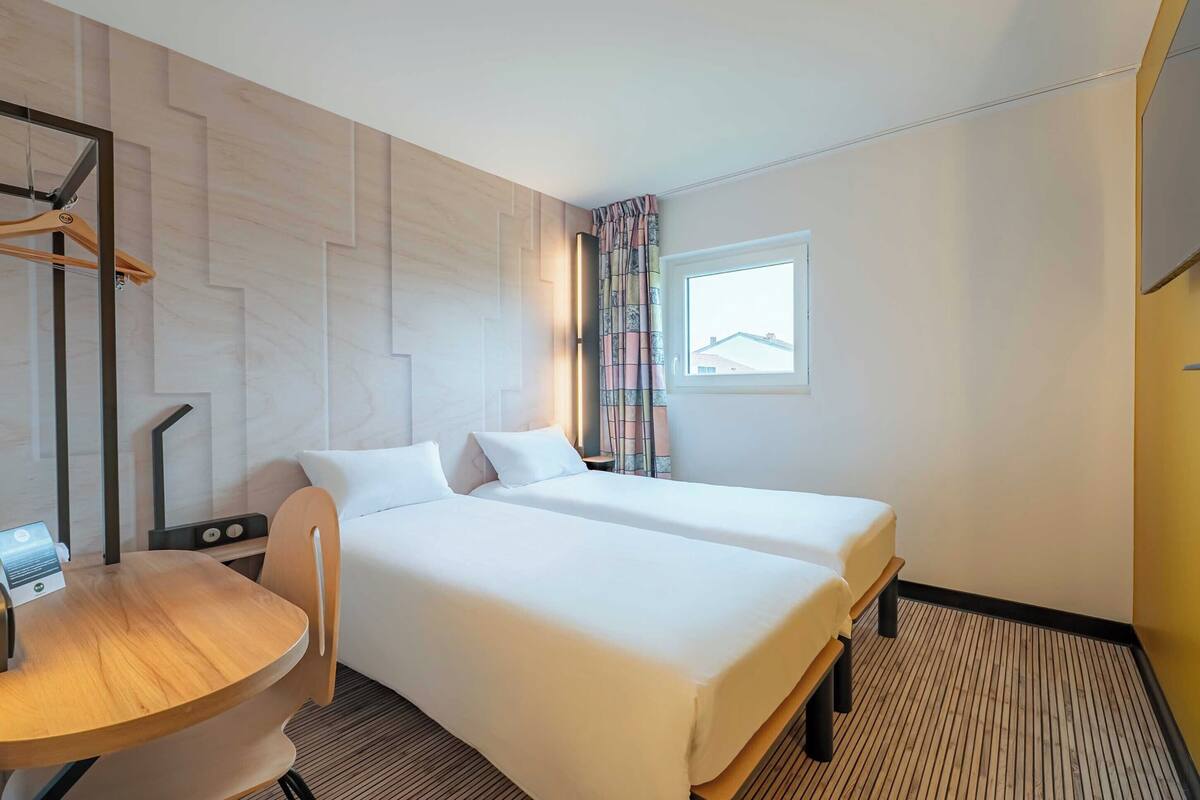 Quarto de hotel com duas camas, uma escrivaninha de madeira, uma cadeira, um cabideiro e uma janela com vista.