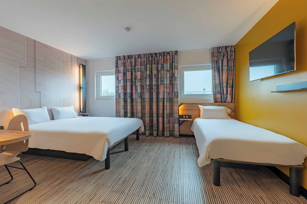 Quarto de hotel com duas camas, uma escrivaninha, uma cadeira, uma televisão e uma janela.