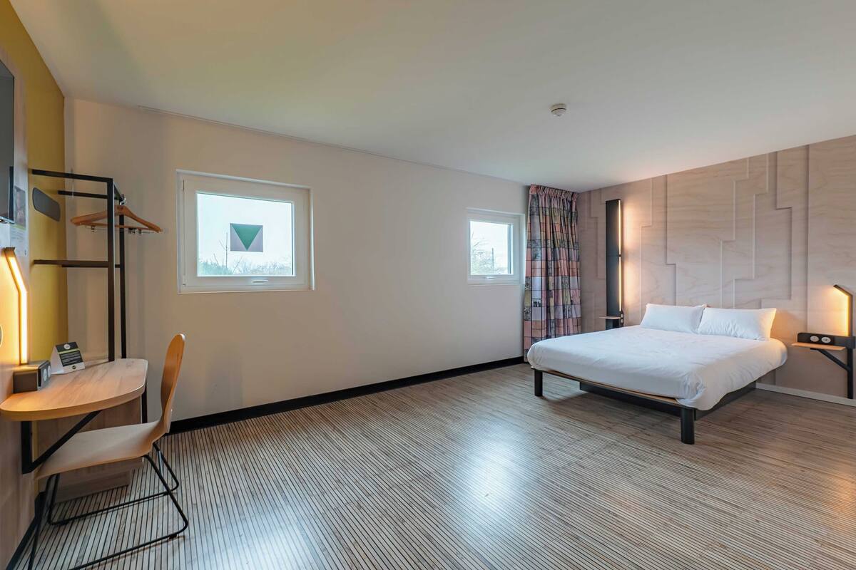 Quarto de hotel com cama, escrivaninha com cadeira, duas janelas e prateleira fixada na parede.