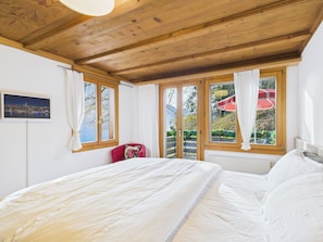 Quarto com teto de madeira, cama com roupa de cama branca, uma cadeira vermelha e um quadro com vista para a cidade.