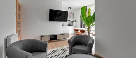 Uma sala de estar moderna com uma TV de tela plana, duas poltronas cinzas, uma mesa de centro redonda e uma planta verde grande.