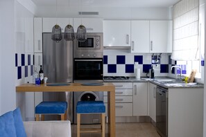 Uma cozinha moderna com armários brancos, uma geladeira de aço inoxidável, forno embutido, lava-louças e uma mesa de madeira com banquetas azuis.