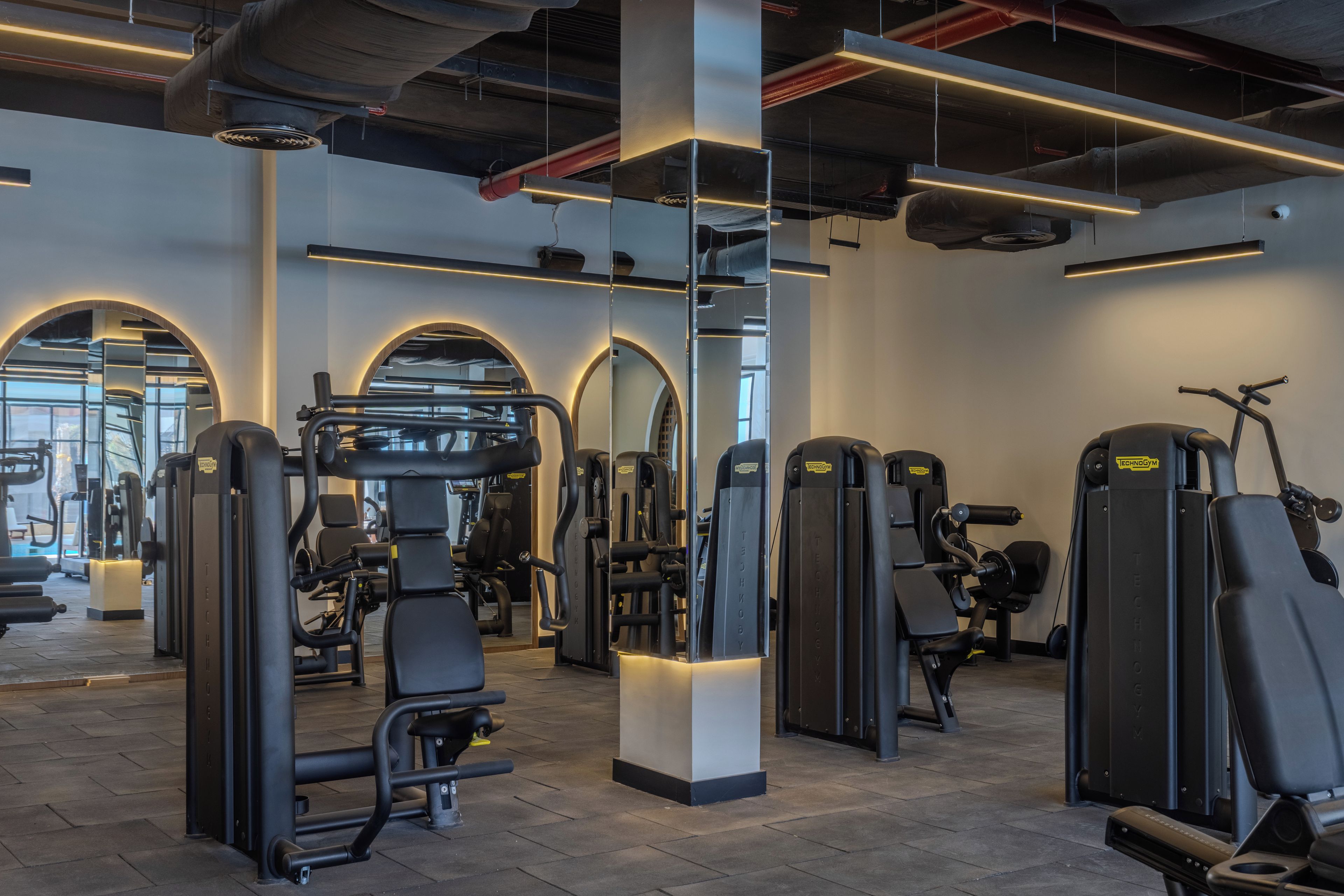 Ginásio com diversos aparelhos de exercício, um espelho na parede e teto com tubulações e iluminação aparentes.
