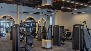 Ginásio com diversos aparelhos de exercício, um espelho na parede e teto com tubulações e iluminação aparentes.