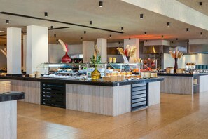 Uma área de buffet com diversas opções de comida, um ponto central de serviço e plantas decorativas.