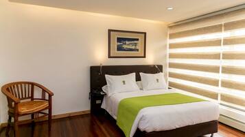 Quarto de hotel com uma cama, uma cadeira, mesa de cabeceira, quadro na parede e janela com persianas.