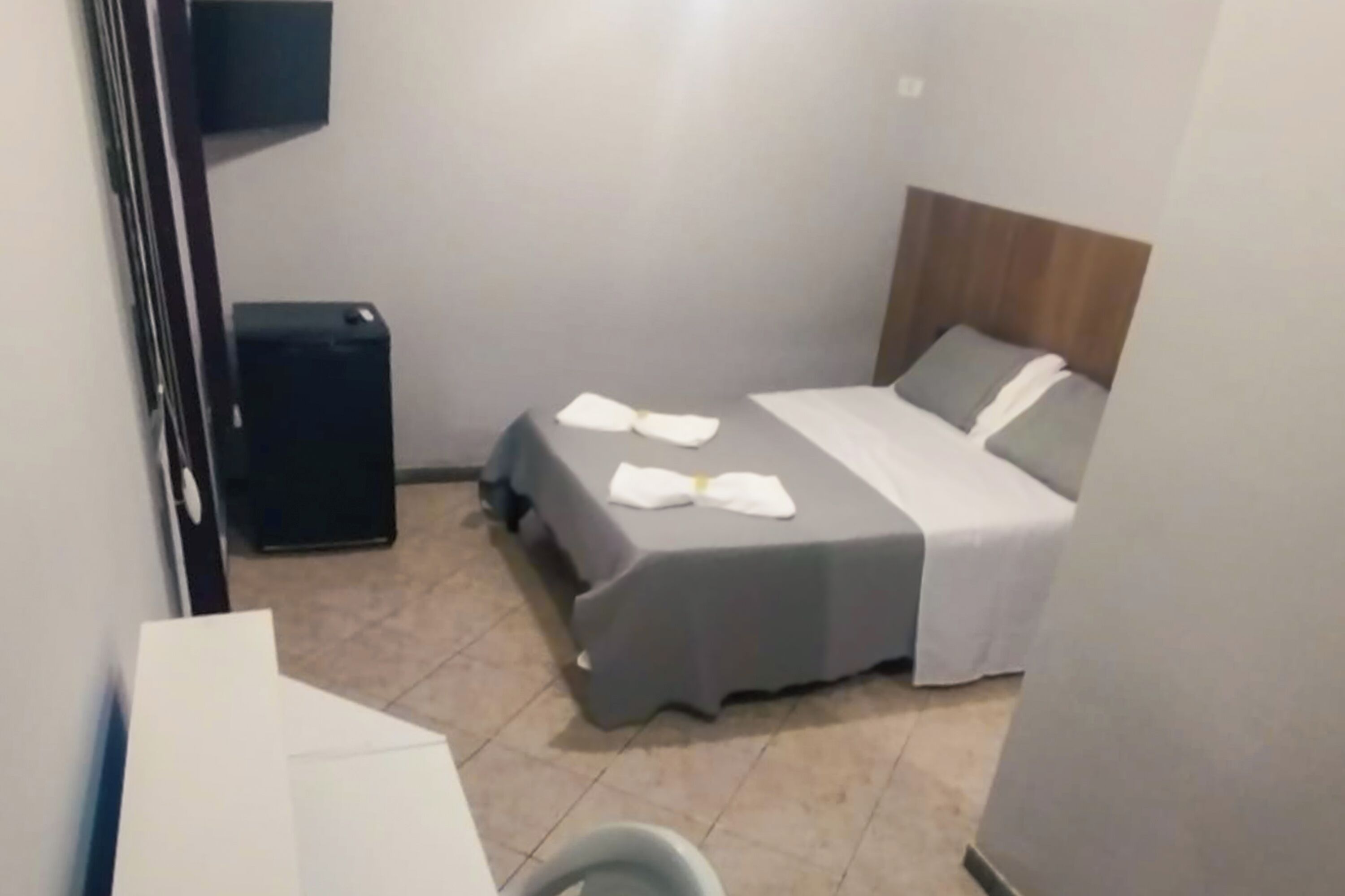 Quarto de hotel pequeno com uma cama, mesa de cabeceira, cadeira, televisão e lixeira.