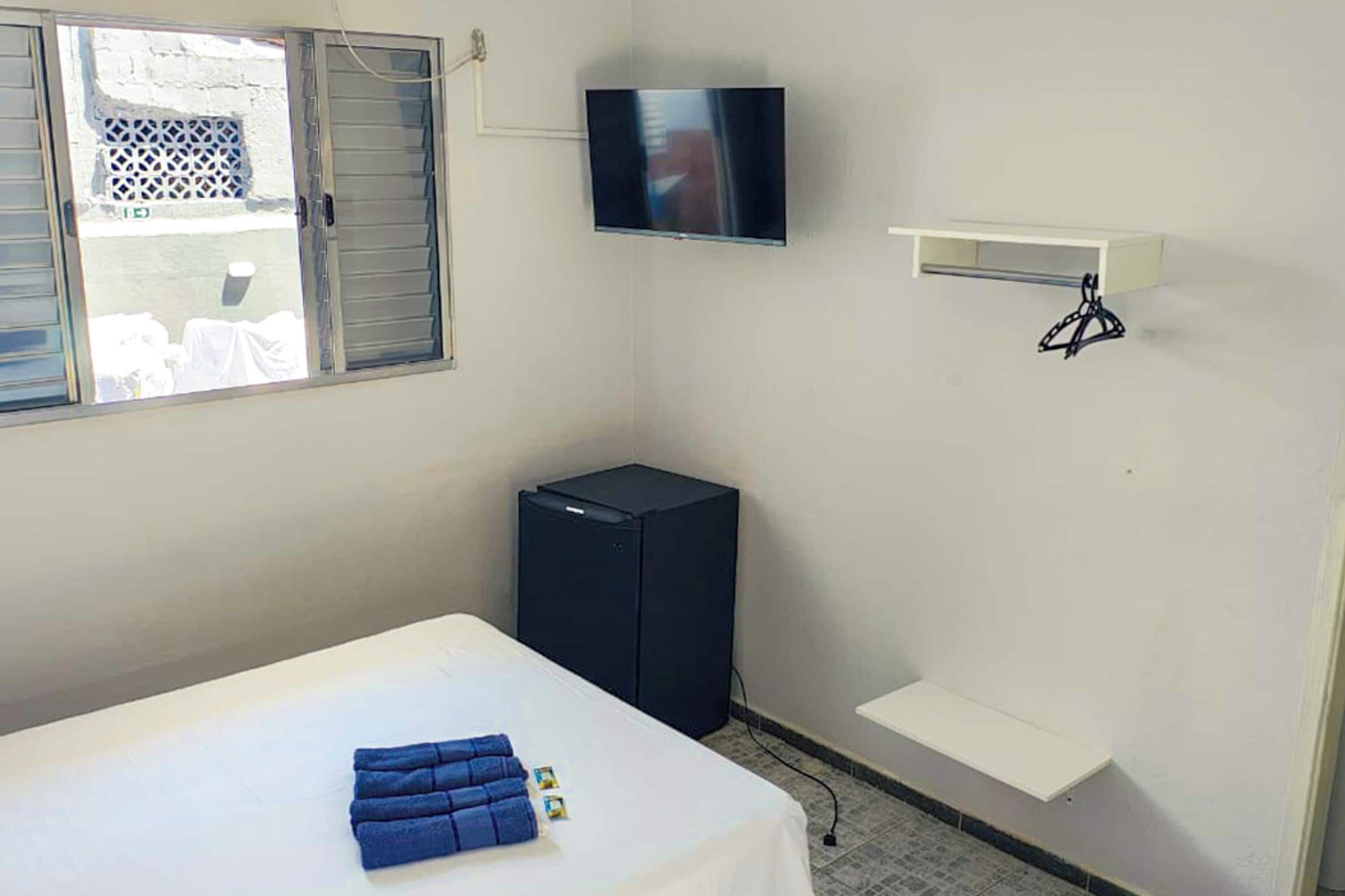Quarto de hotel com cama, televisão fixada na parede, um pequeno armário preto e janela com persianas.