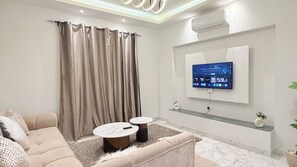 Uma sala de estar moderna com um sofá, duas mesinhas de centro, uma TV fixada na parede e um aparelho de ar condicionado.