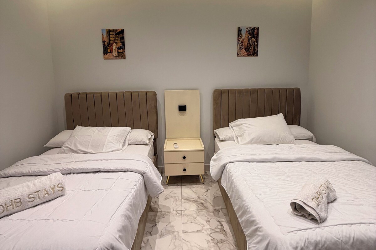 Quarto de hotel com duas camas, uma mesa de cabeceira, uma mesinha e uma luminária no teto.