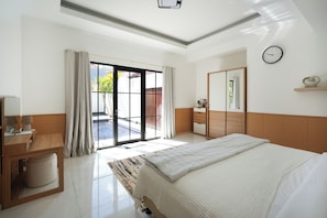 Quarto de hotel com cama, escrivaninha, espelho, relógio e porta de vidro corrediça para área externa.