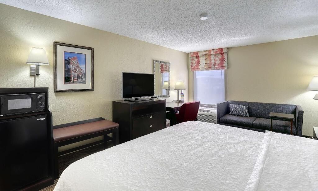 Quarto de hotel com uma cama grande, televisão, escrivaninha, cadeira, sofá e janela com cortinas.