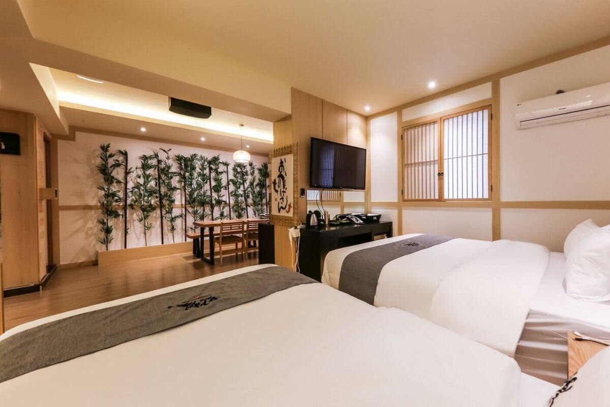 Quarto de hotel com cama, escrivaninha, televisão, ar condicionado e área de refeições com plantas.