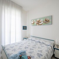 Quarto com cama, mesa de cabeceira, tomada na parede e janela com cortinas.