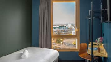 Quarto de hotel com cama, uma cadeira, uma mesinha com um vaso e uma janela com vista para um aeroporto.
