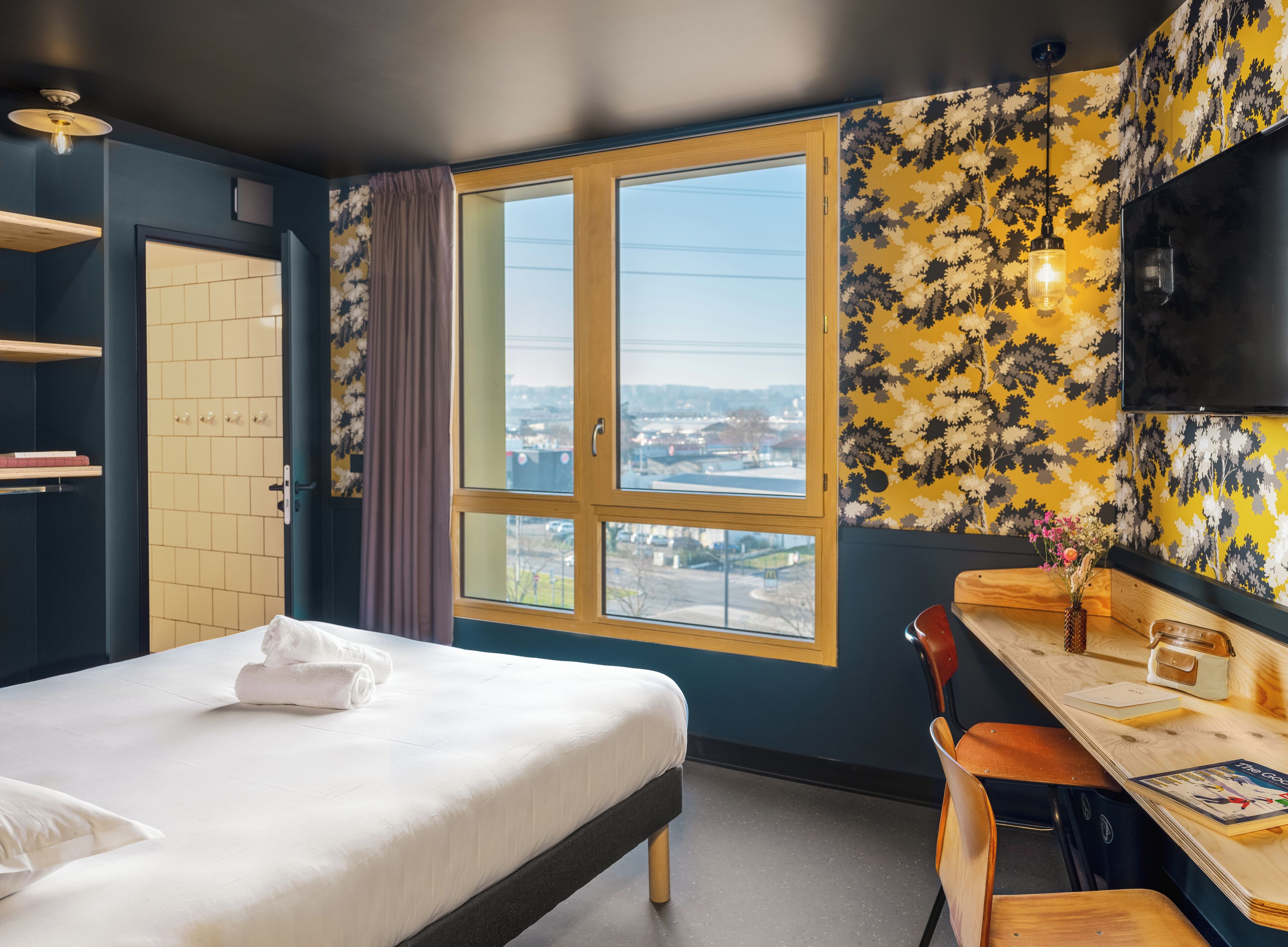 Quarto de hotel com cama, escrivaninha, cadeira, televisão e janela com vista para a cidade.